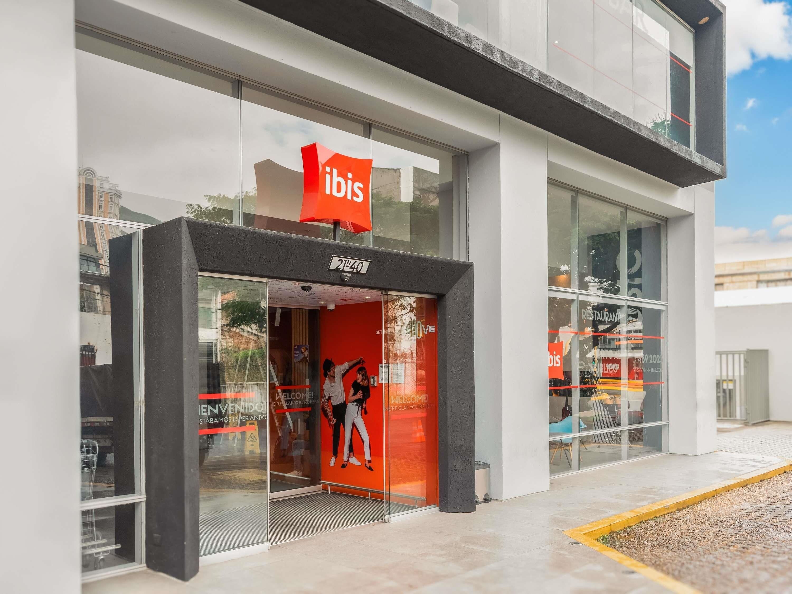 Um edifício moderno com fachada de vidro e um grande letreiro vermelho da 'ibis', além de um cartaz de boas-vindas.