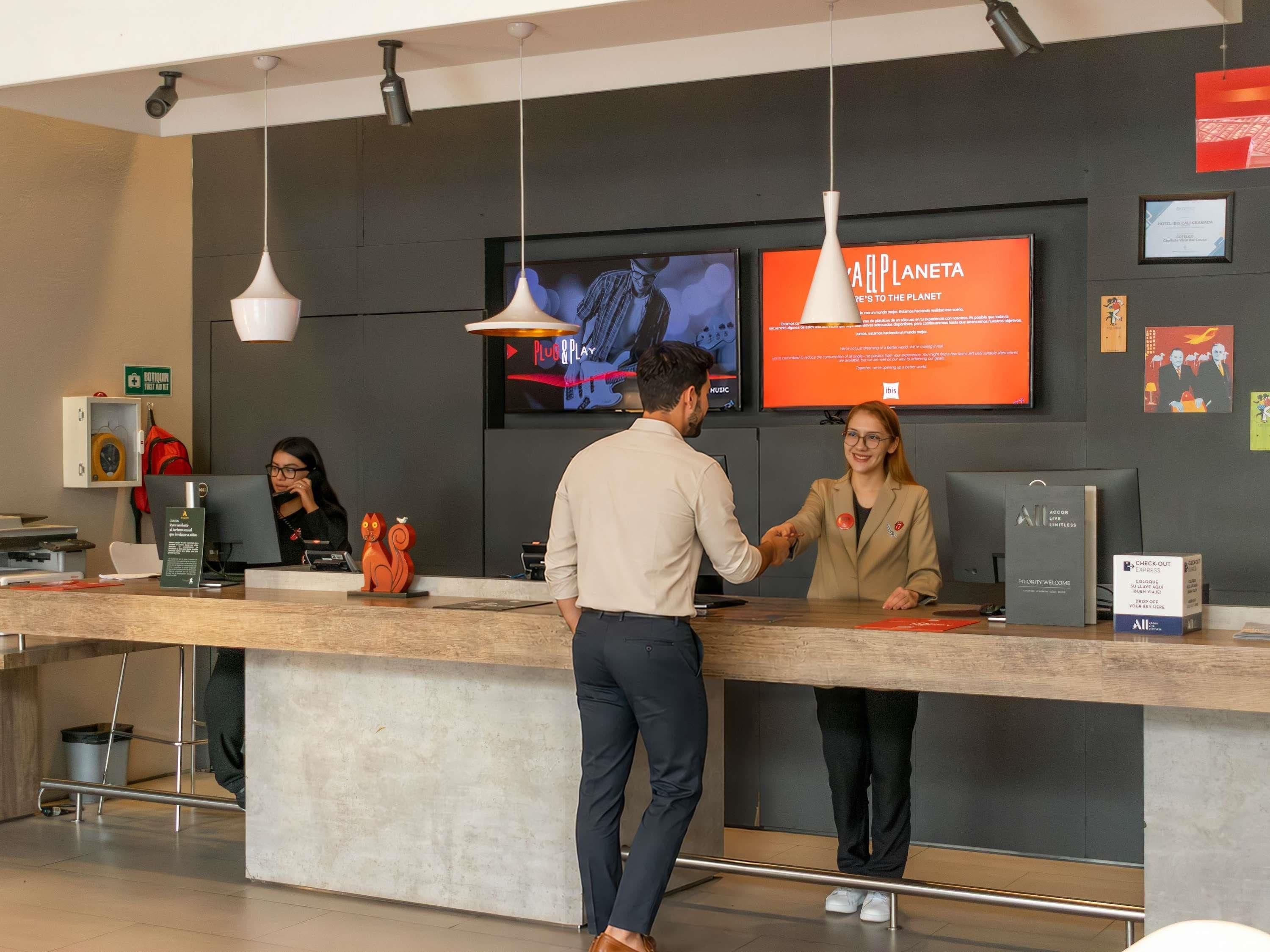Bancada de recepção com tela digital exibindo um vídeo, sinalização para serviço de check-out rápido e uma obra de arte emoldurada na parede.