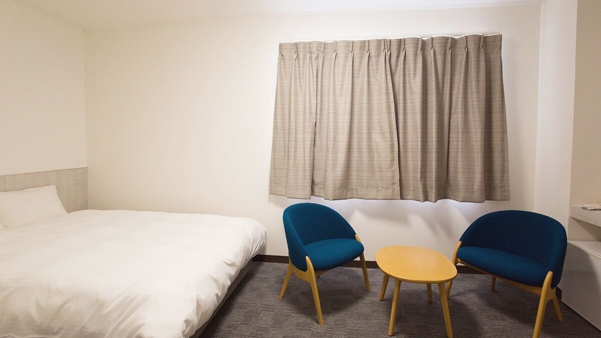Quarto de hotel com uma cama, duas cadeiras azuis, uma mesa redonda pequena e uma janela com cortinas.