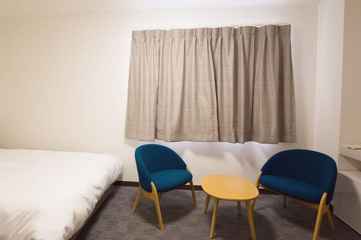 Quarto de hotel com uma cama, duas cadeiras azuis, uma mesa redonda pequena e uma janela com cortinas.