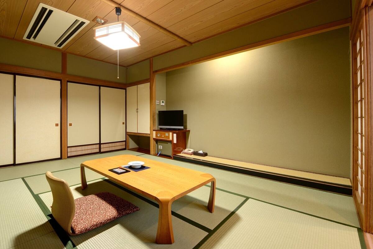 Um quarto japonês tradicional com tatames, uma mesa de madeira baixa, uma cadeira estofada, portas de correr e uma televisão.
