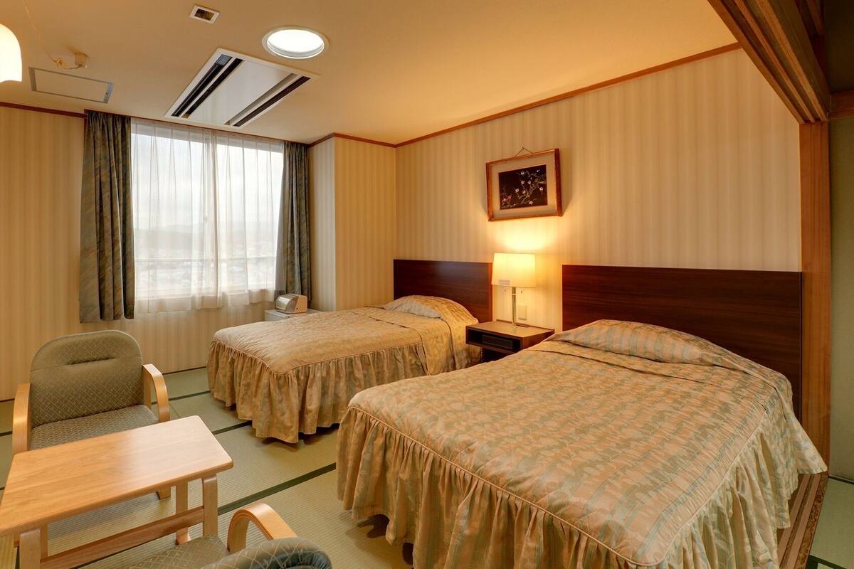 Quarto de hotel com duas camas, uma escrivaninha, uma cadeira, mesa de cabeceira, abajur e um quadro na parede.