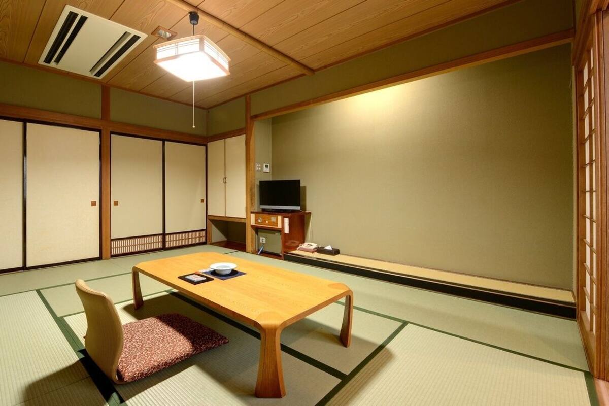 Um quarto japonês tradicional com tatames, uma mesa de madeira baixa, uma cadeira, portas de correr e uma televisão.