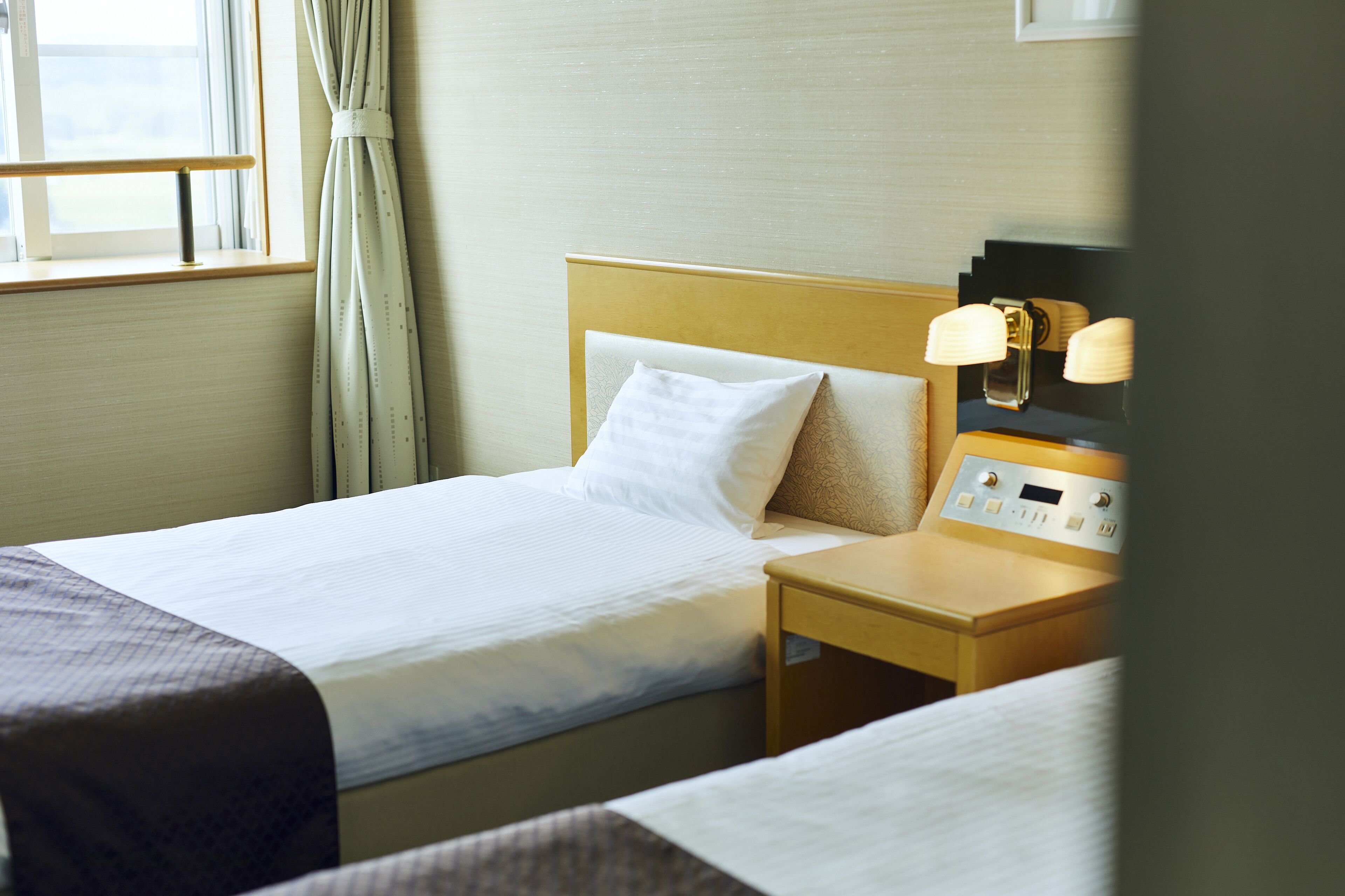 Quarto de hotel com cama, mesa de cabeceira, luminária e janela com cortinas.