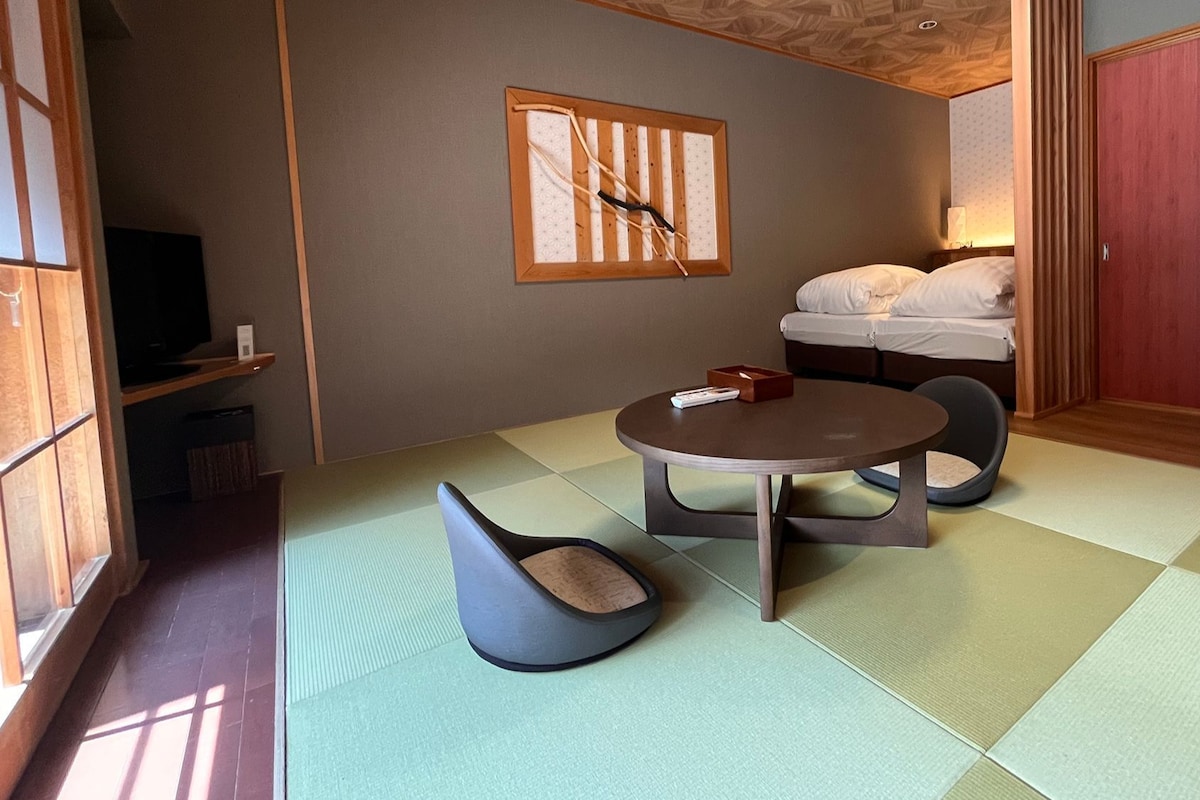 Quarto com tatames, uma mesa baixa com cadeiras, uma cama com roupa de cama branca e uma TV fixada na parede.