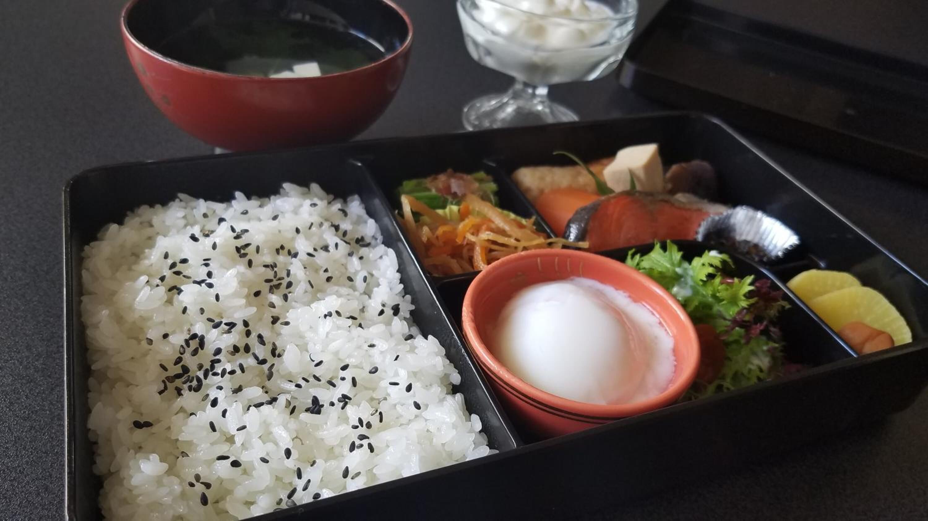 Uma caixa bento com arroz branco, legumes, peixe e uma pequena tigela de sopa.