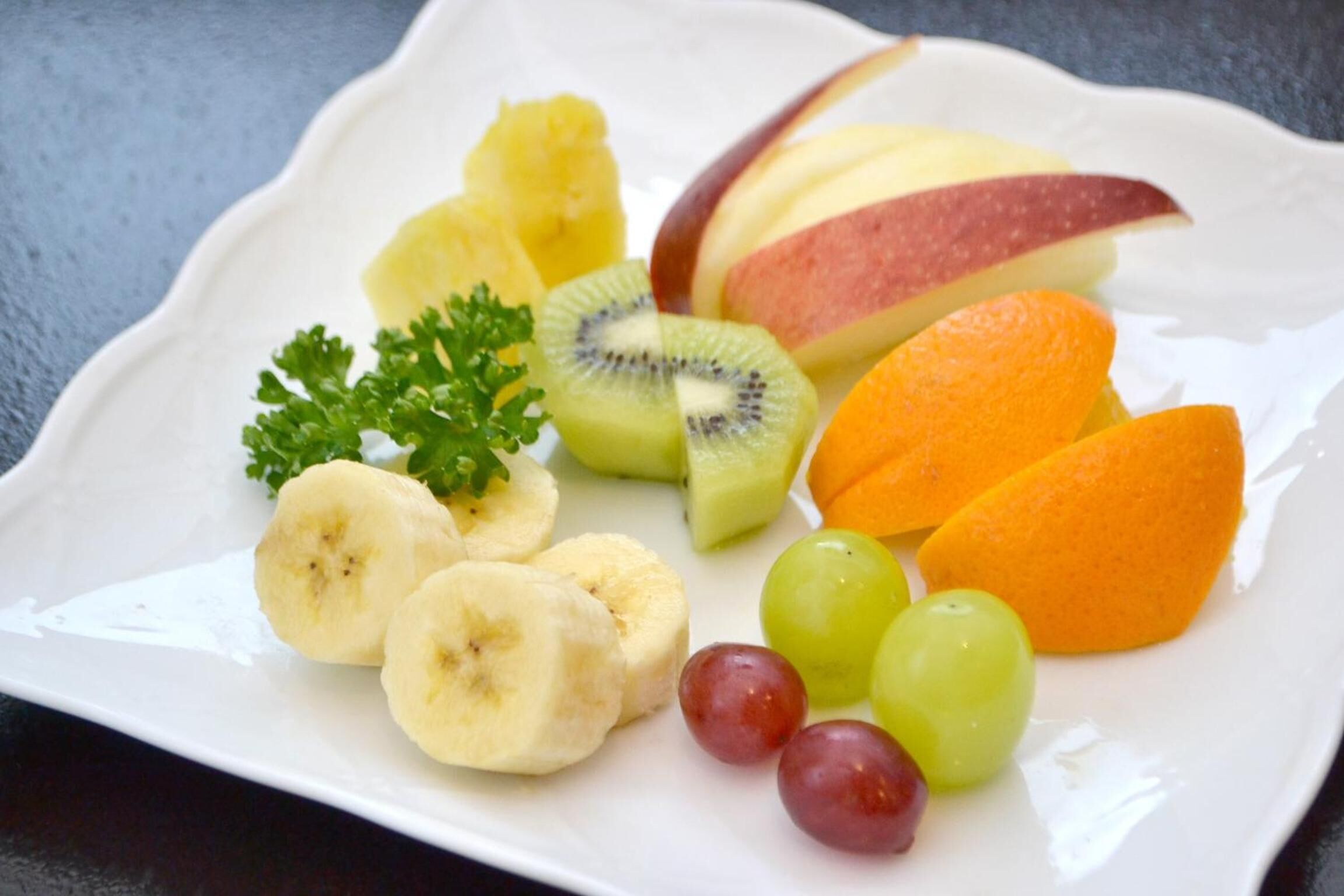 Um prato branco com frutas fatiadas, incluindo bananas, kiwi, maçãs, laranjas e uvas.