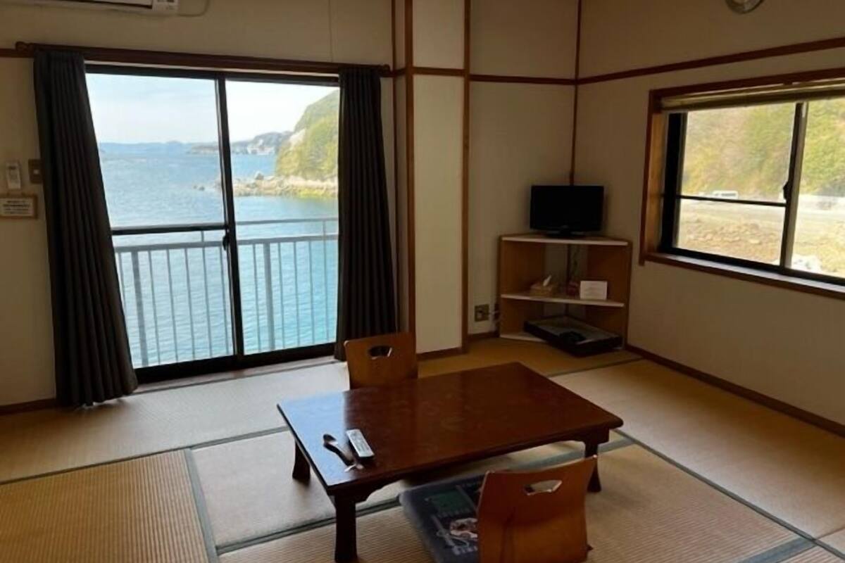 Quarto com piso de tatame, uma mesa de madeira, televisão sobre um suporte e vista para o mar através de uma grande janela.