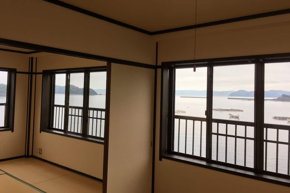 Quarto com piso de tatame, janelas amplas com molduras pretas e vista para um corpo d'água com montanhas ao longe.