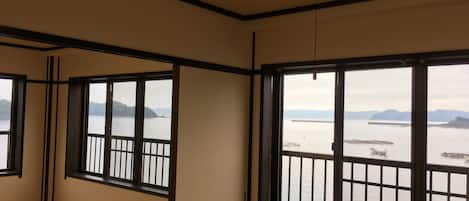 Quarto com piso de tatame, janelas amplas com molduras pretas e vista para um corpo d'água com montanhas ao longe.