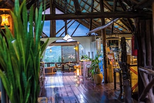 Um interior rústico com vigas de madeira, uma janela grande com vista para uma paisagem montanhosa e um espaço de estar com mesas e cadeiras.