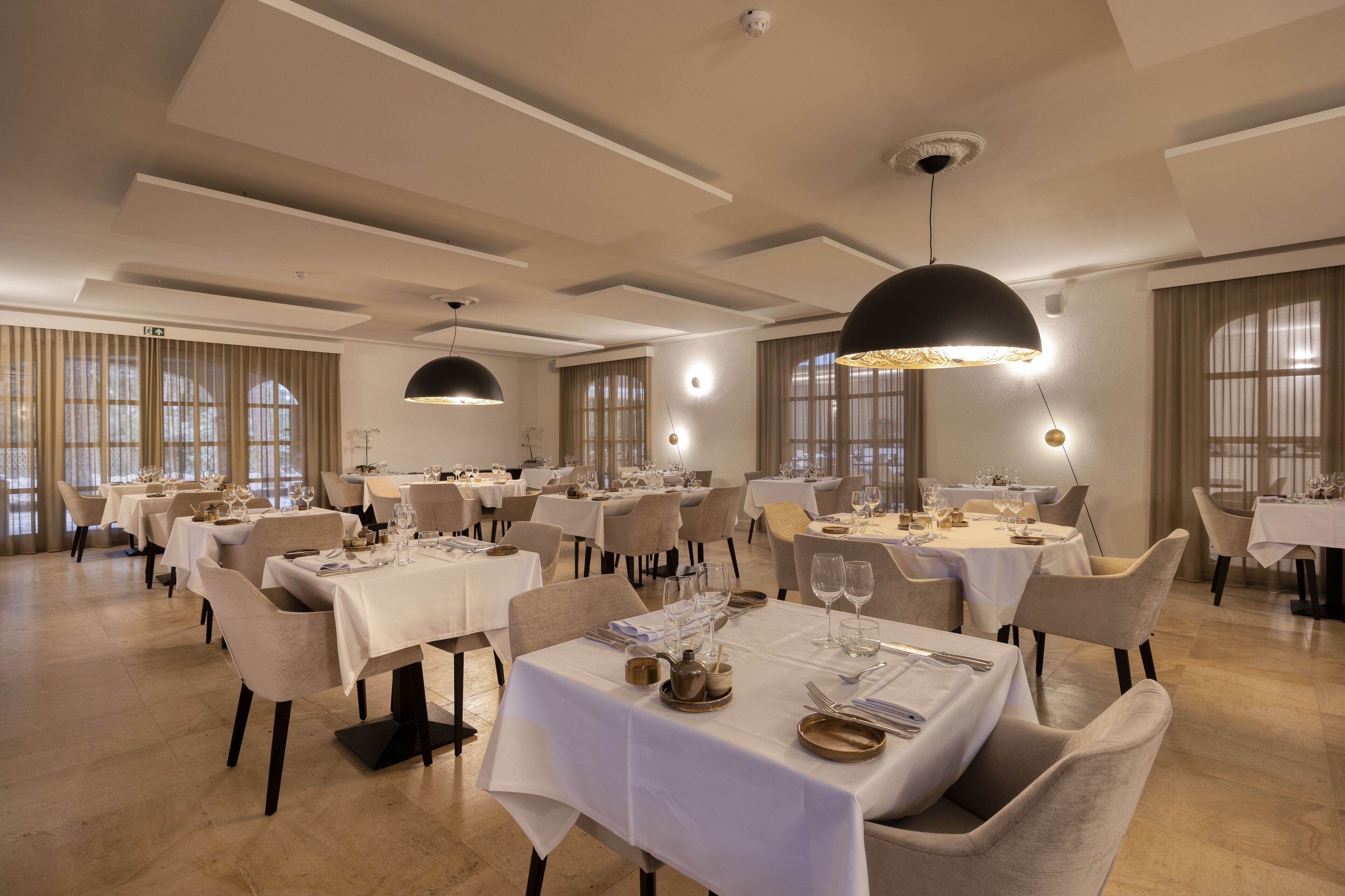 Uma ampla sala de jantar com várias mesas preparadas para refeições, apresentando um design de teto moderno e grandes janelas.