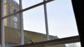 Vista pela janela de um edifício com uma torre do relógio, uma porta azul e um espaço gramado.