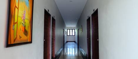 Um corredor longo e estreito, com uma pintura grande na parede esquerda, portas de madeira em ambos os lados e chão de azulejos.