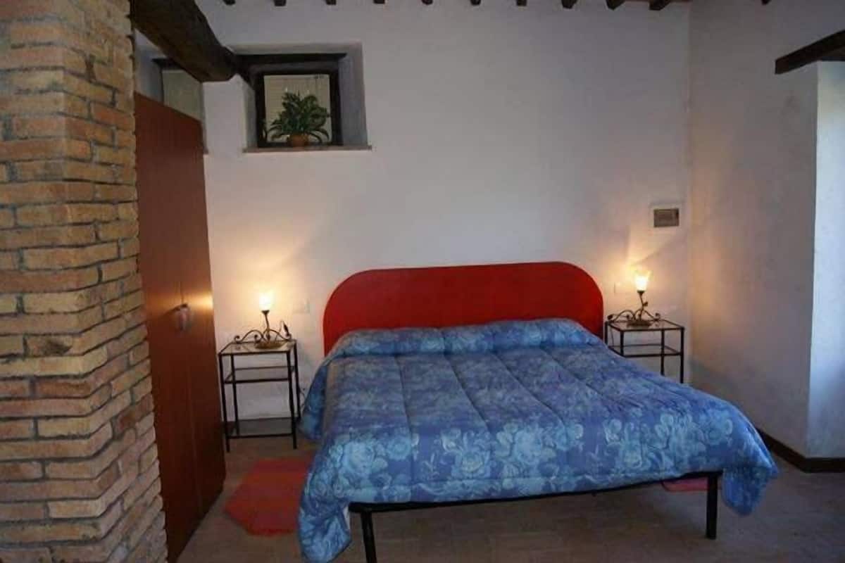 Quarto com parede de tijolos, cabeceira vermelha, colcha florida azul e duas mesas de cabeceira com abajures.