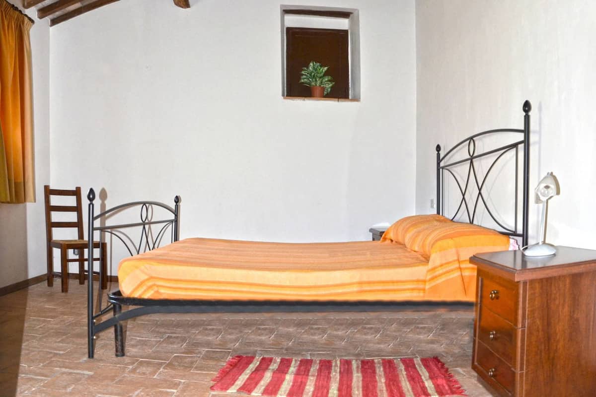 Quarto com cama, cadeira de madeira, mesa de cabeceira pequena, luminária e janela com planta em vaso.
