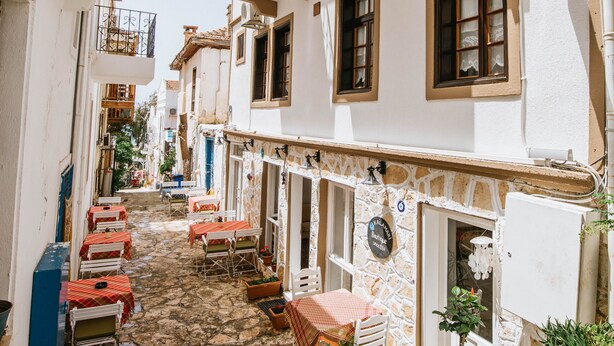Uma rua estreita com edifícios de ambos os lados, uma bandeira e mesas ao ar livre.