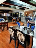 Uma área de refeições com mesas de madeira e cadeiras listradas, uma parede de tijolos e um espaço de bar com cardápio em lousa.