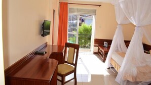 Quarto de hotel com secretária de madeira, uma cadeira, cama com dossel, televisão e varanda com vista para área verde.