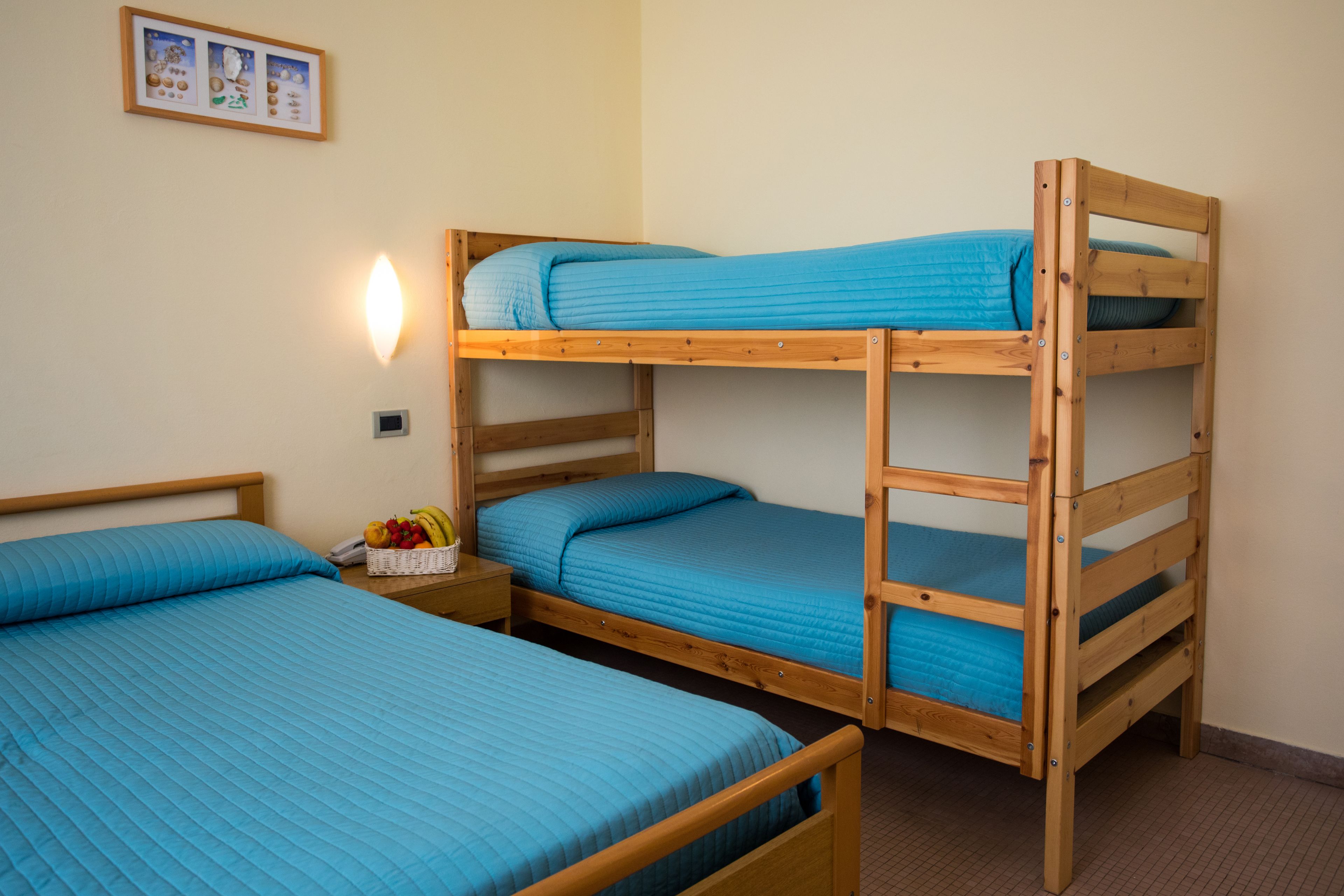 Quarto com duas camas beliche, cada uma com roupa de cama azul, uma mesa de cabeceira com uma cesta de frutas e uma luminária na parede.