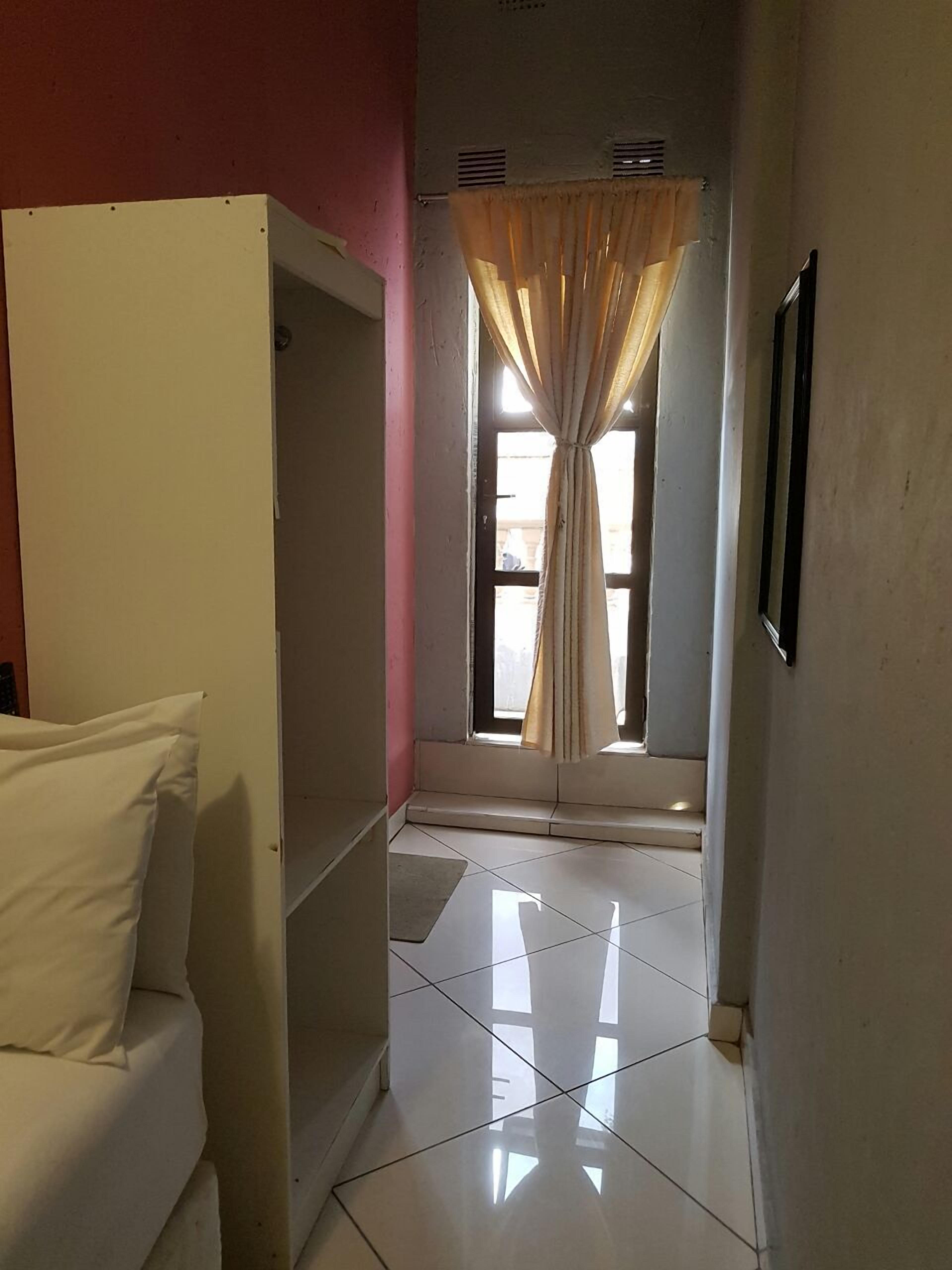 Um corredor estreito com chão de azulejos brancos, um guarda-roupa branco entreaberto e uma janela com cortinas bege.