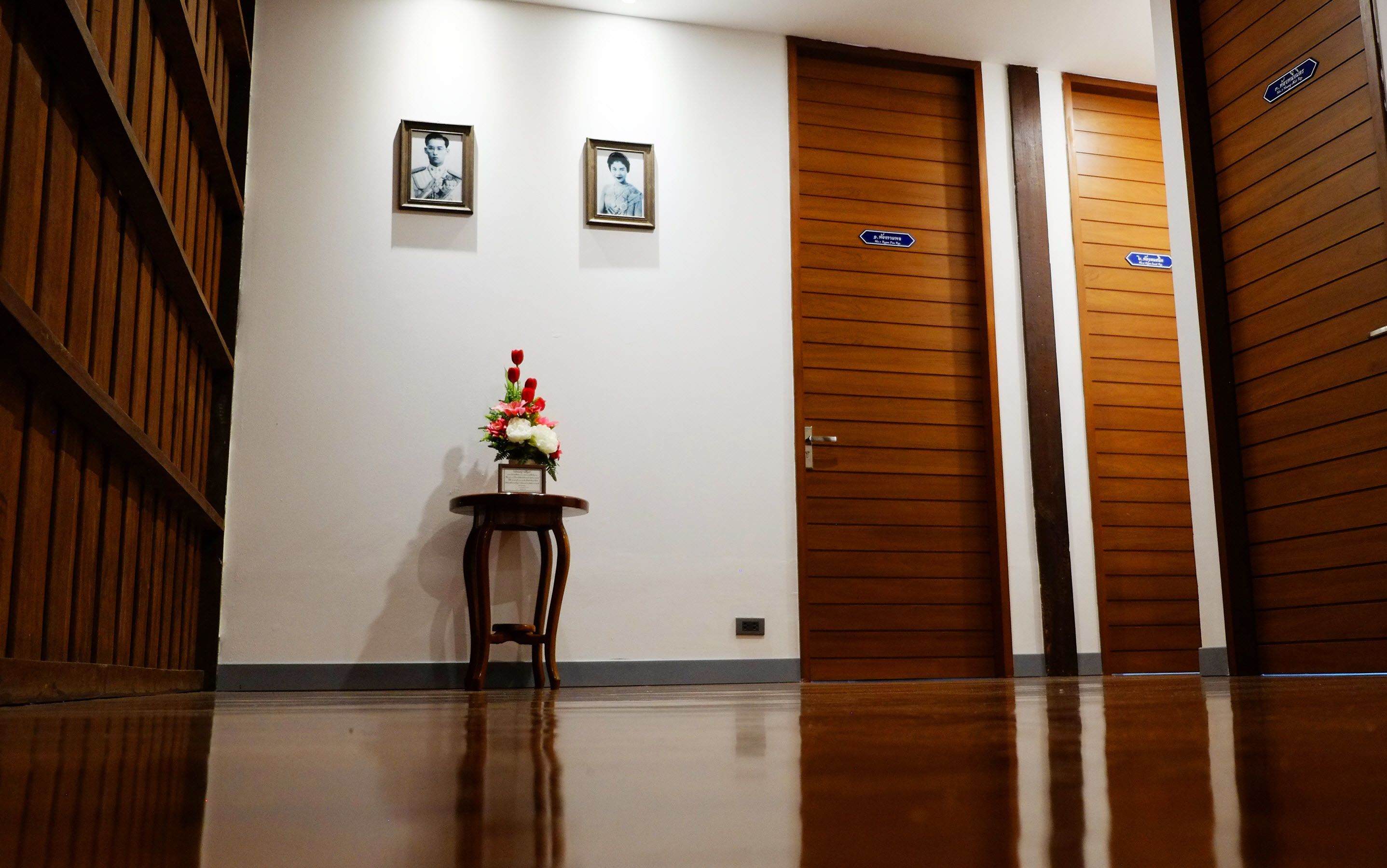 Um corredor com portas de madeira, uma pequena mesa com um arranjo floral e duas molduras na parede.