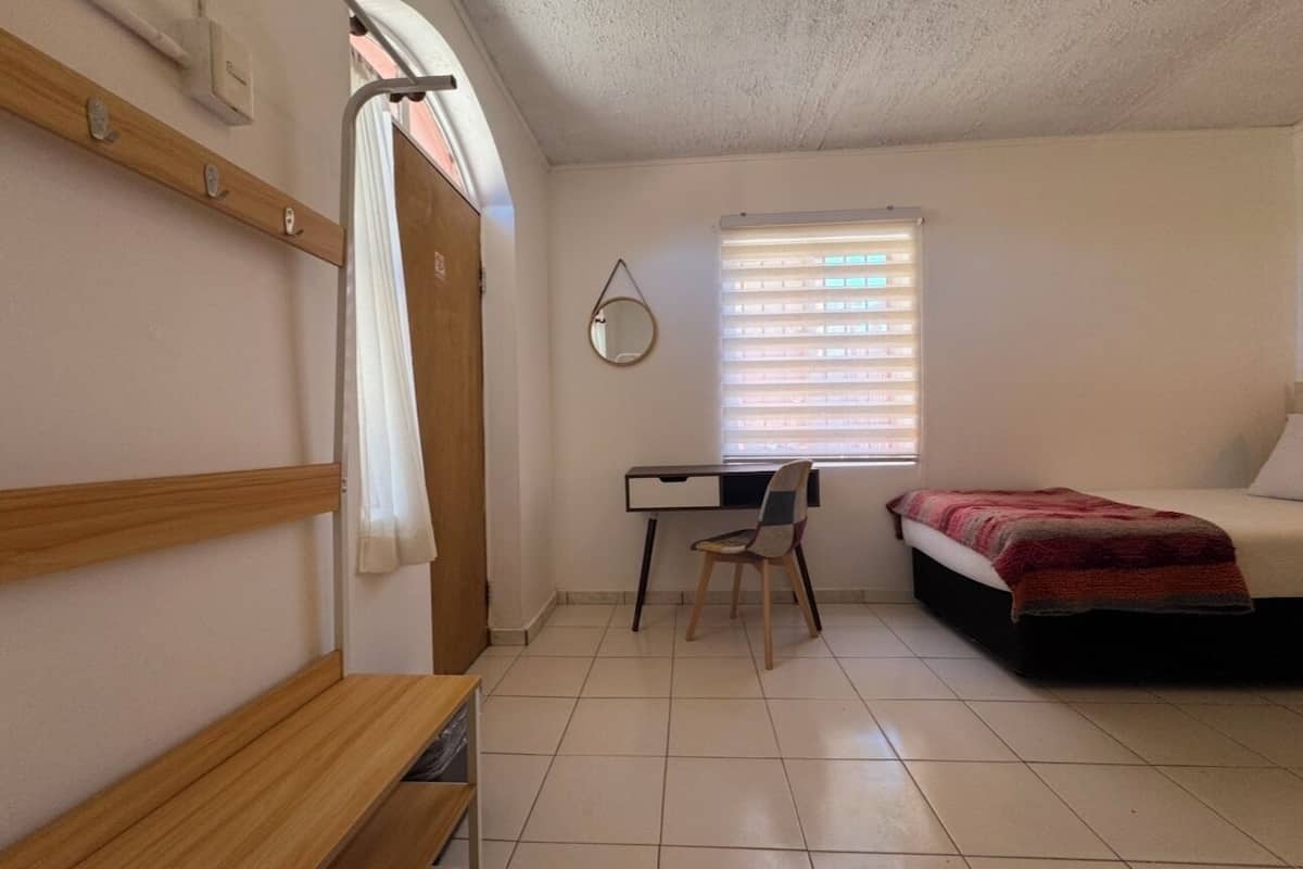 Um quarto de estudante com cama, escrivaninha, cadeira e janela com persianas.