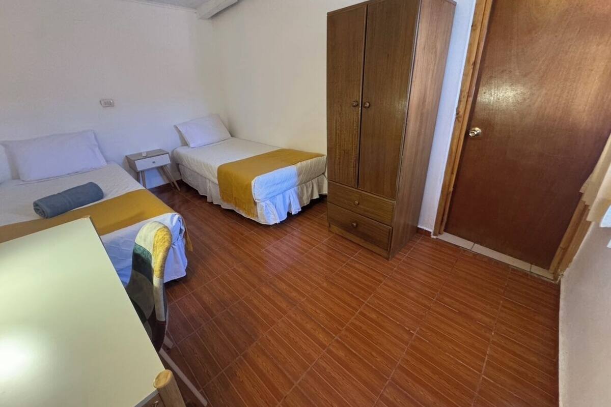 Quarto pequeno e arrumado com duas camas individuais, um guarda-roupa de madeira, uma porta de madeira e uma mesa pequena.