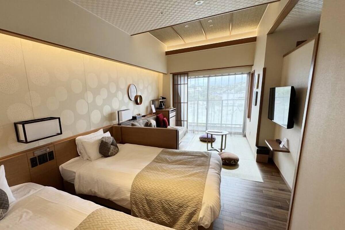 Quarto de hotel com duas camas, uma escrivaninha, uma televisão e uma janela grande.