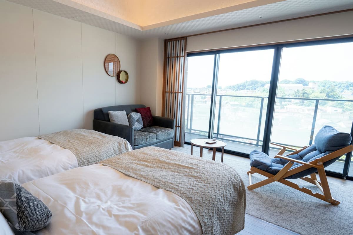 Quarto de hotel com duas camas, um sofá, uma cadeira, uma mesinha e amplas janelas com vista.