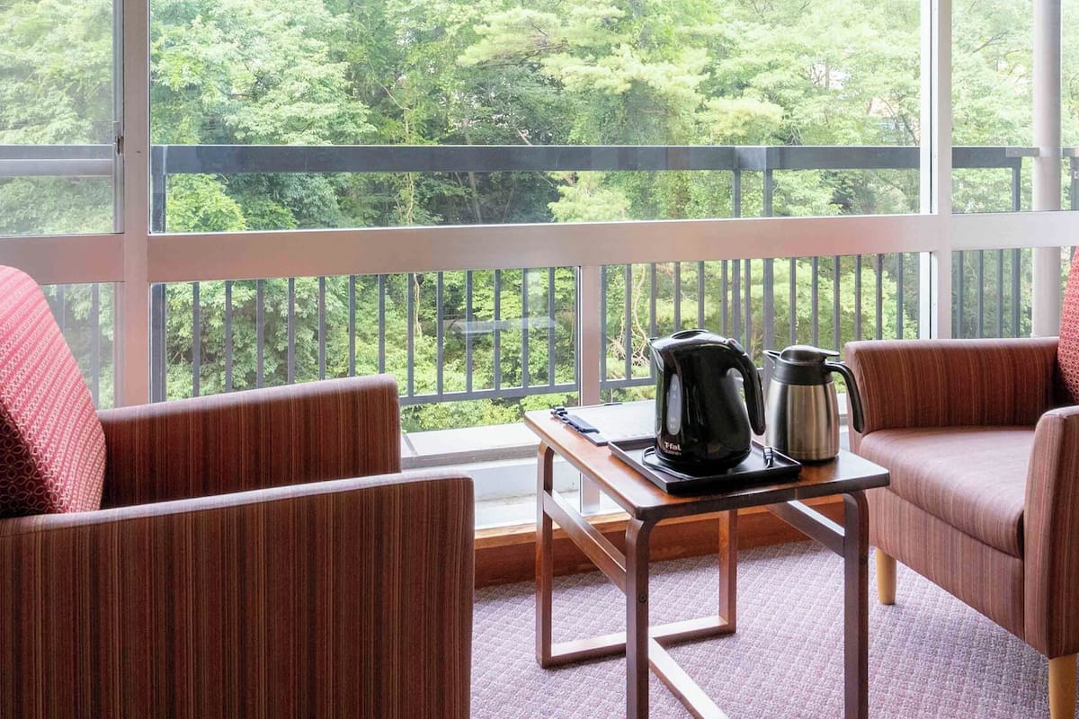 Uma varanda com duas poltronas, uma mesa com cafeteira e chaleira, e vista para uma floresta.