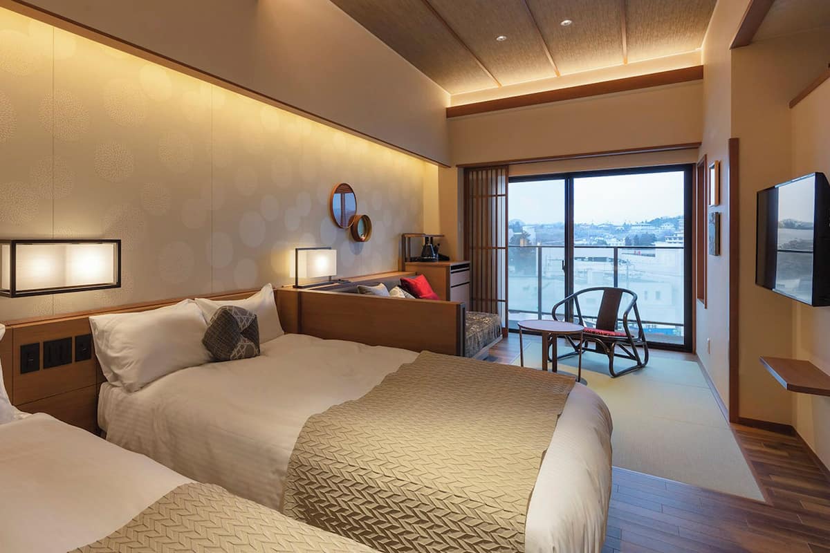 Quarto de hotel com duas camas, uma escrivaninha, uma cadeira, televisão e varanda com vista.