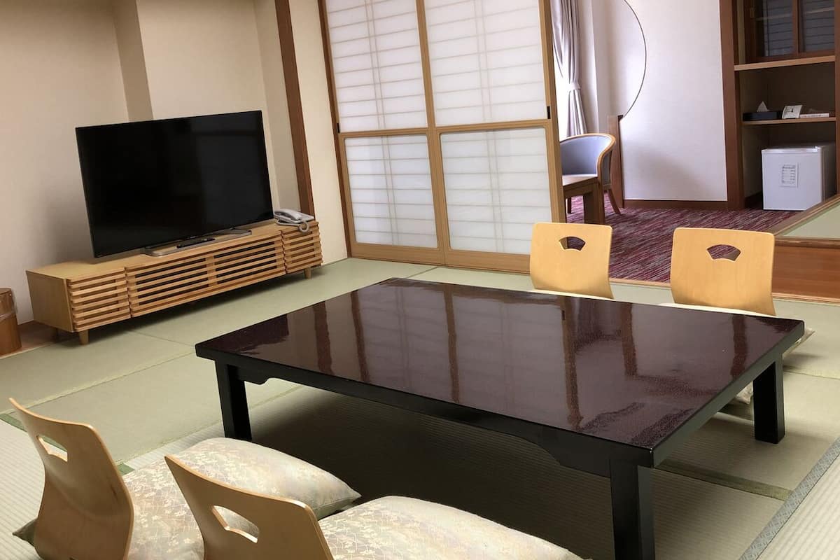 Um quarto tradicional japonês com piso de tatame, uma mesa baixa, duas cadeiras de madeira, uma televisão e uma porta corrediça com biombos shoji.
