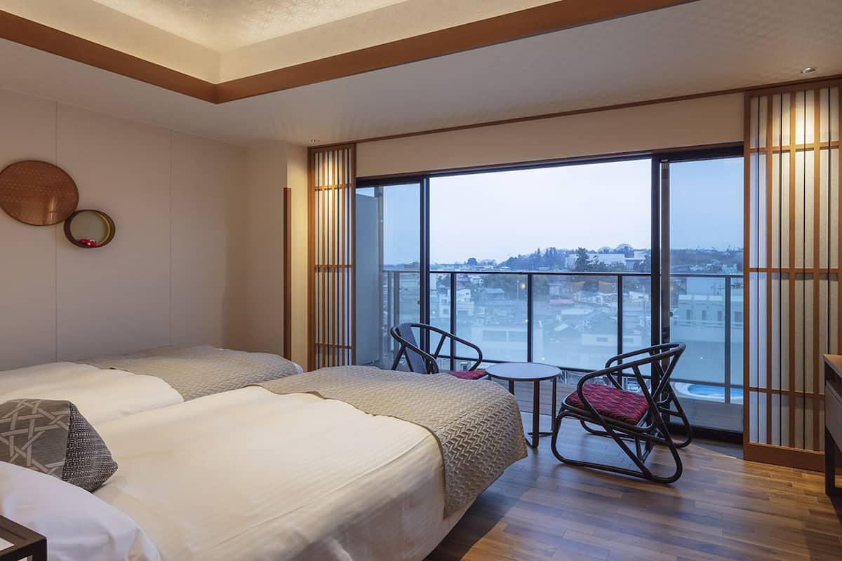 Quarto de hotel com duas camas, uma mesinha, uma cadeira, televisão e uma janela grande com vista para a cidade.