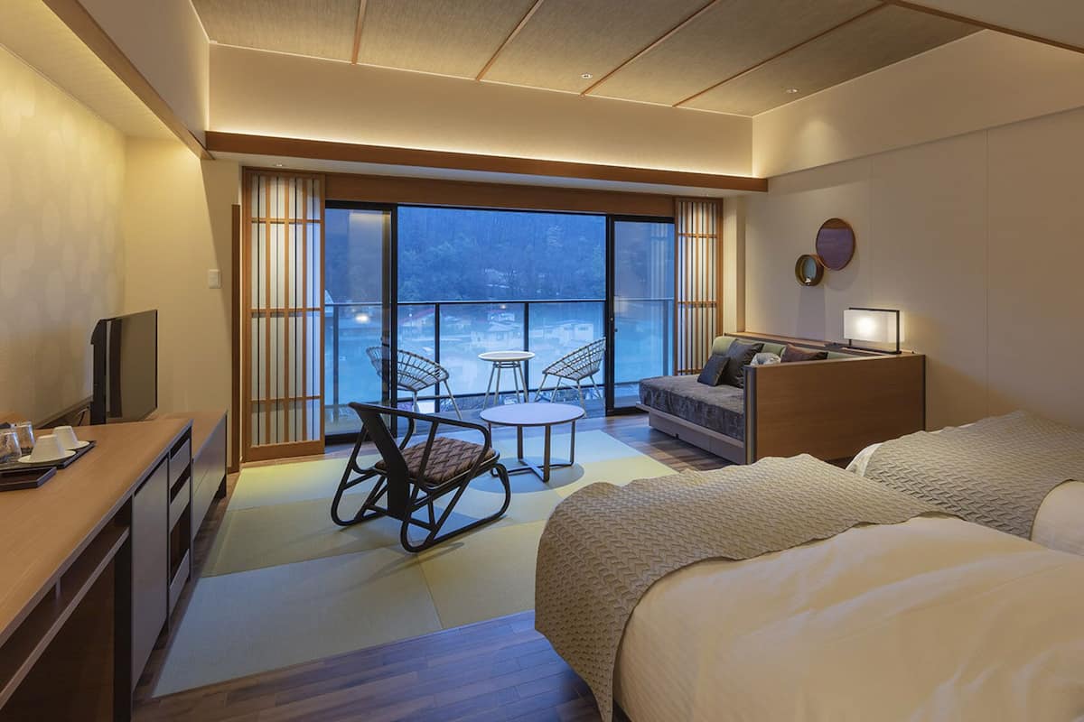 Quarto de hotel com cama, escrivaninha, cadeira, mesa pequena e vista através de amplas janelas.