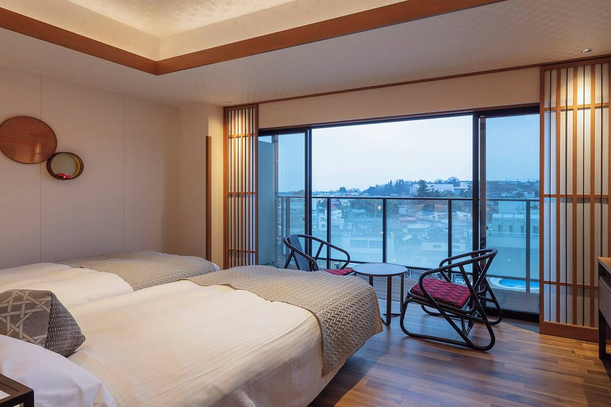 Quarto de hotel com duas camas, uma mesinha com cadeiras, televisão e uma janela grande com vista para a cidade.