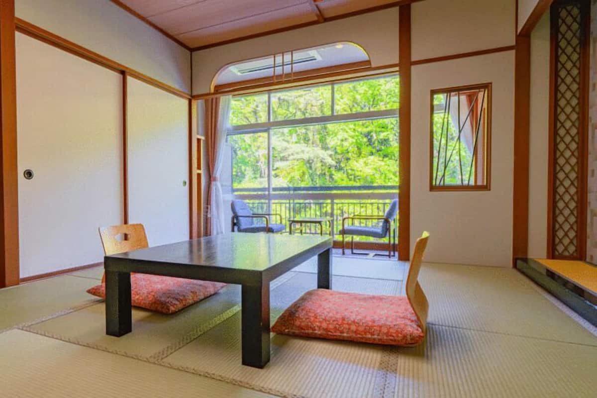 Um quarto japonês tradicional com tatames, uma mesa baixa e vista para o verde através de grandes janelas.
