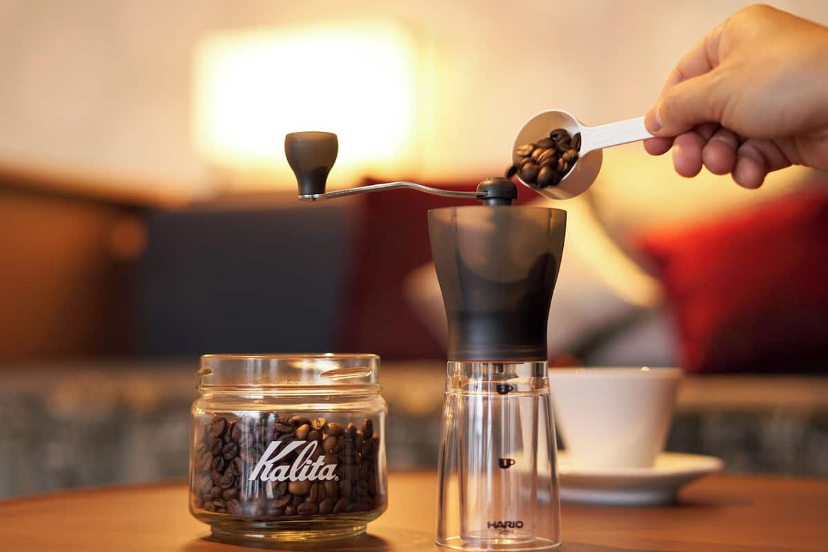 Uma mão moendo café com um moedor, com um pote de grãos de café Kalita e um moedor transparente sobre uma mesa.