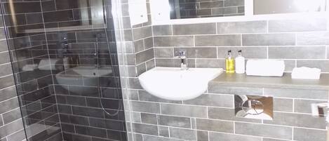 Casa de banho com chuveiro, lavatório e sanita, com paredes em azulejos escuros e pavimento com padrão.