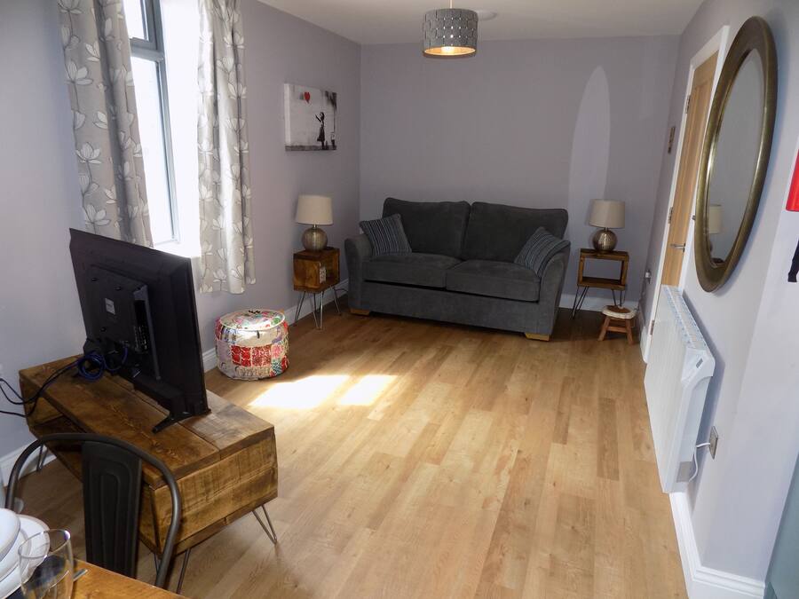 Uma sala de estar com um sofá cinzento, uma mesa de centro de madeira, uma televisão e um abajur sobre uma mesinha lateral.