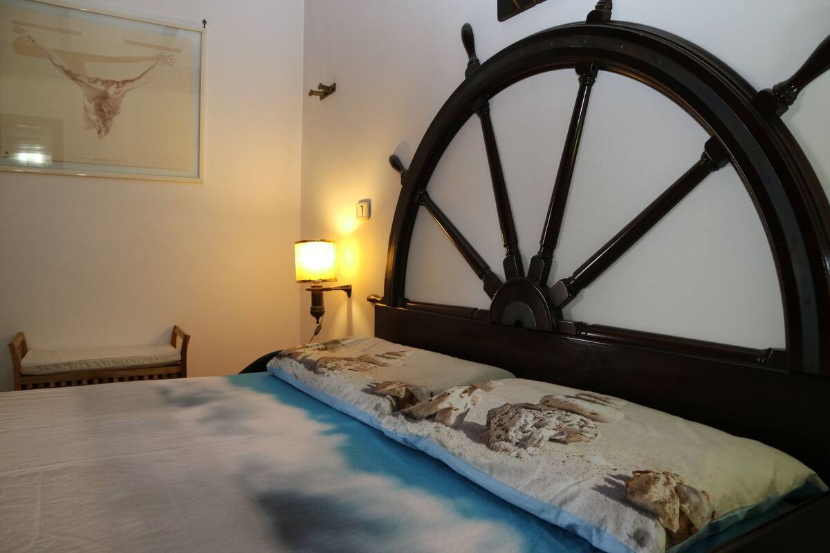 Uma cama com cabeceira de madeira, abajur na mesa de cabeceira e um quadro pendurado na parede.