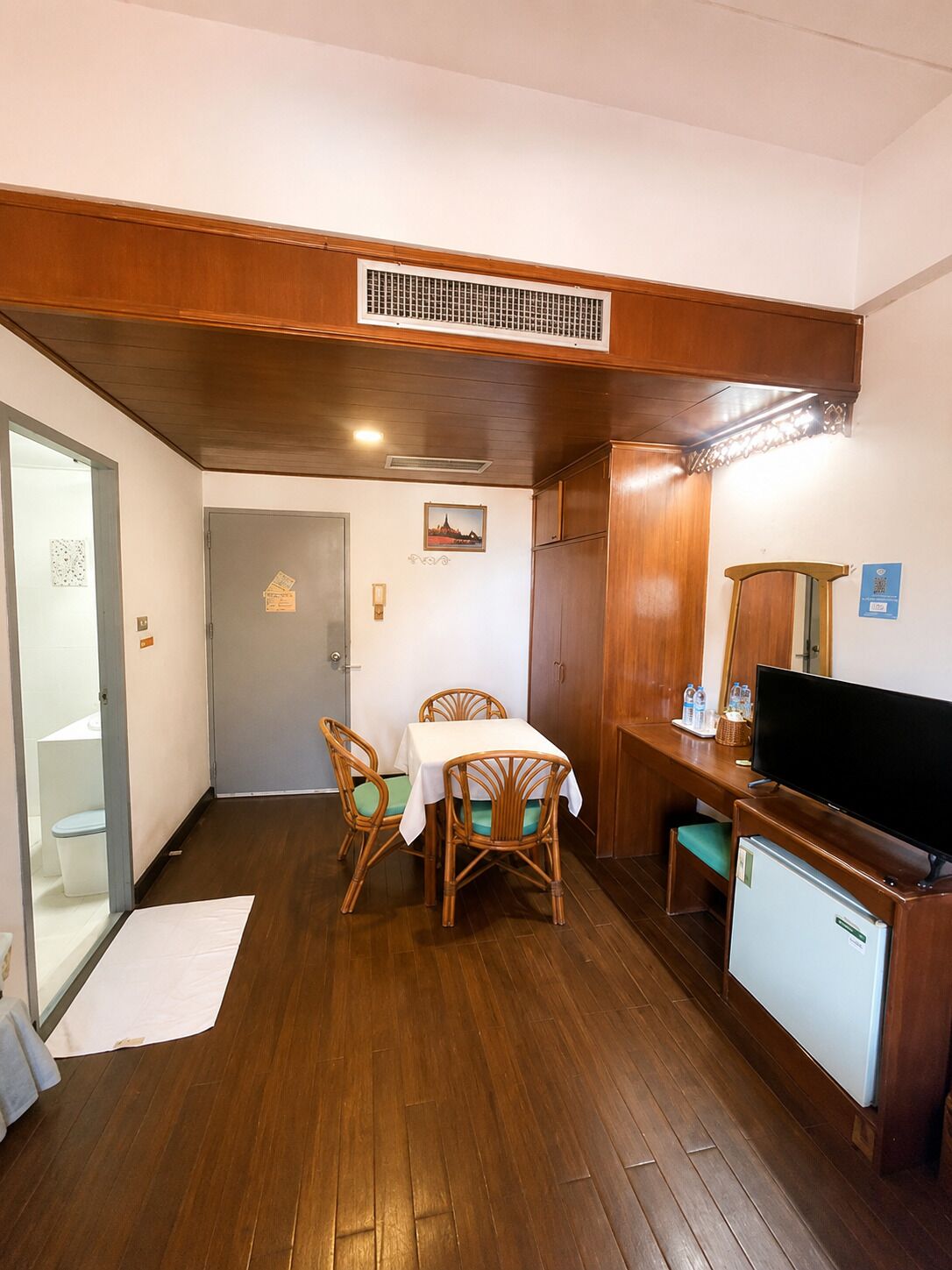 Um quarto com piso de madeira, uma mesa com cadeiras, uma televisão, um pequeno frigorífico e uma porta que dá para outro quarto.