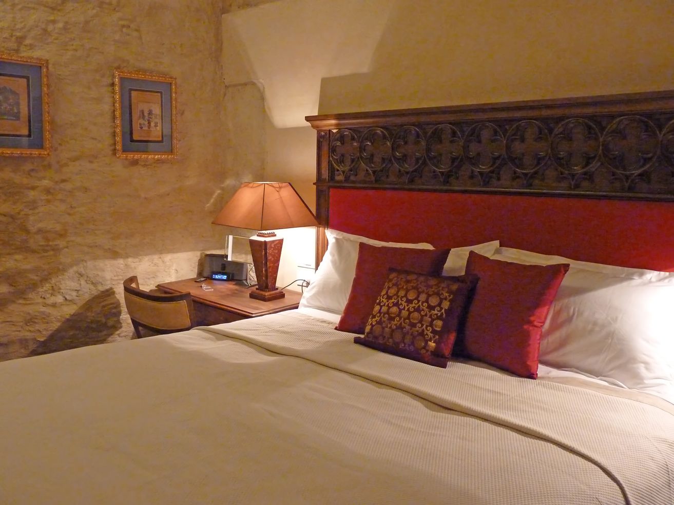 Cama bem arrumada com almofadas vermelhas e estampadas, mesa de cabeceira de madeira com abajur e quadros nas paredes.