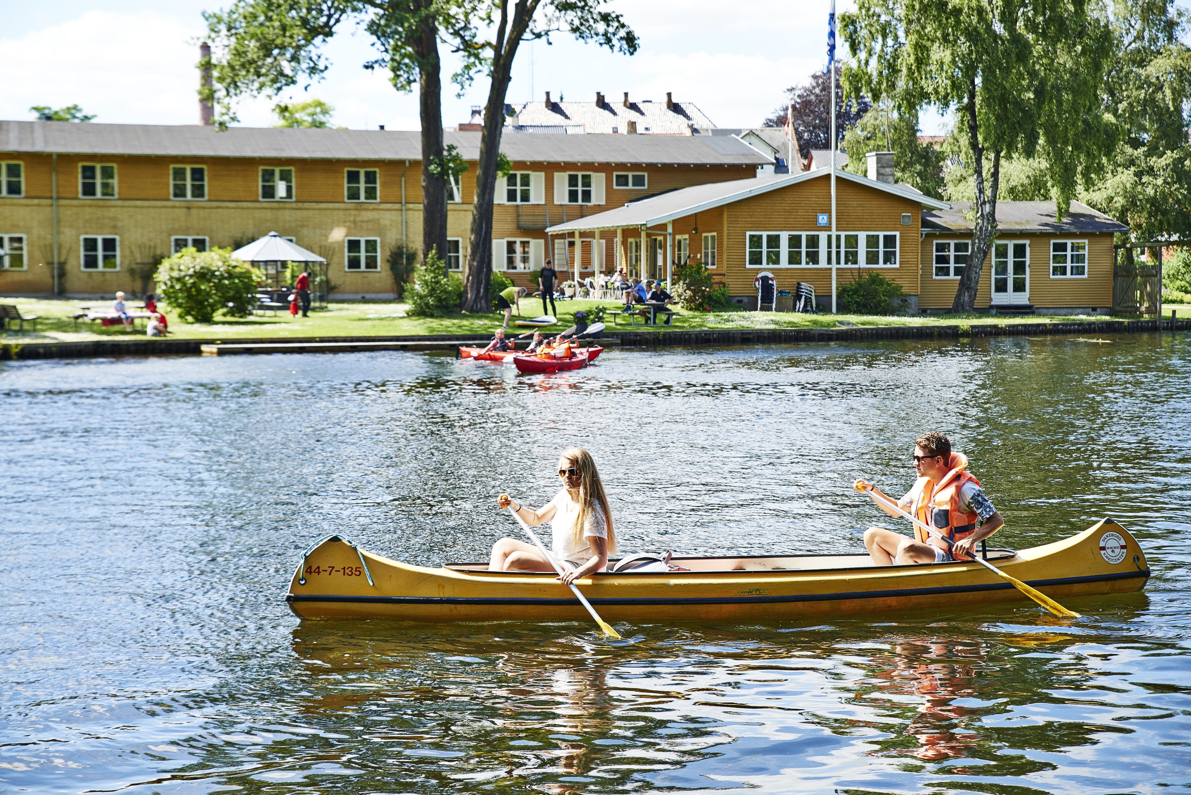 Um lago calmo com um canoa amarela em primeiro plano e um edifício com um gramado ao fundo.