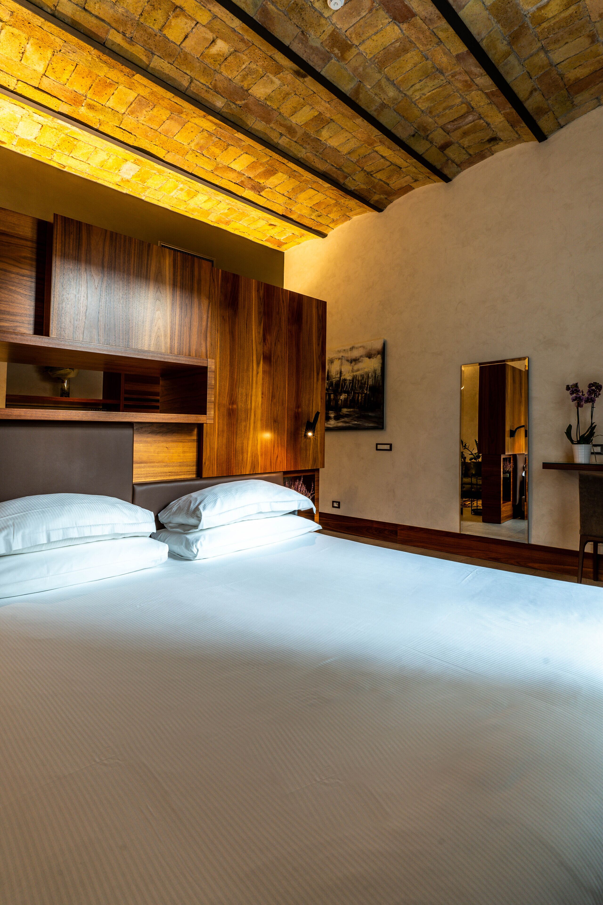 Quarto de hotel com uma cama grande, cabeceira de madeira, estantes embutidas, um espelho e um quadro na parede.