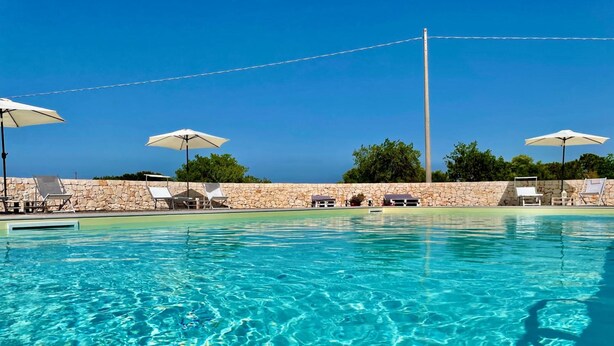 Piscina azul límpida com espreguiçadeiras e guarda-sóis, rodeada por um muro de pedra e árvores.
