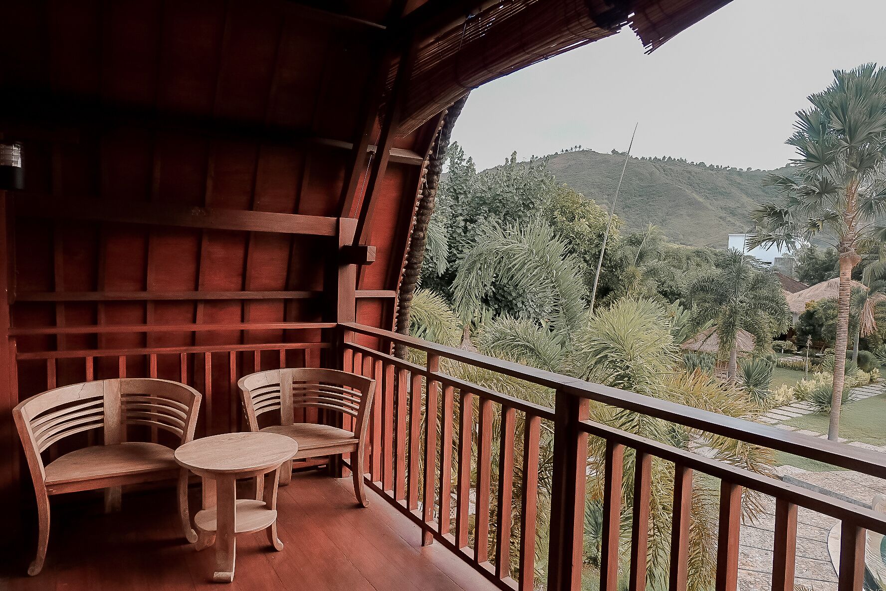 Varanda de madeira com duas cadeiras e uma mesa pequena, com vista para uma paisagem exuberante com árvores e colinas.