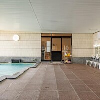 Uma área de piscina com piso de azulejos, porta de vidro que dá para um quarto, e uma fileira de cadeiras brancas à direita.