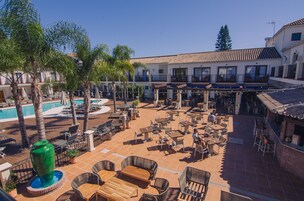 Uma área de piscina no terraço com espreguiçadeiras e mesas, cercada por palmeiras e um edifício com telhado de azulejos.