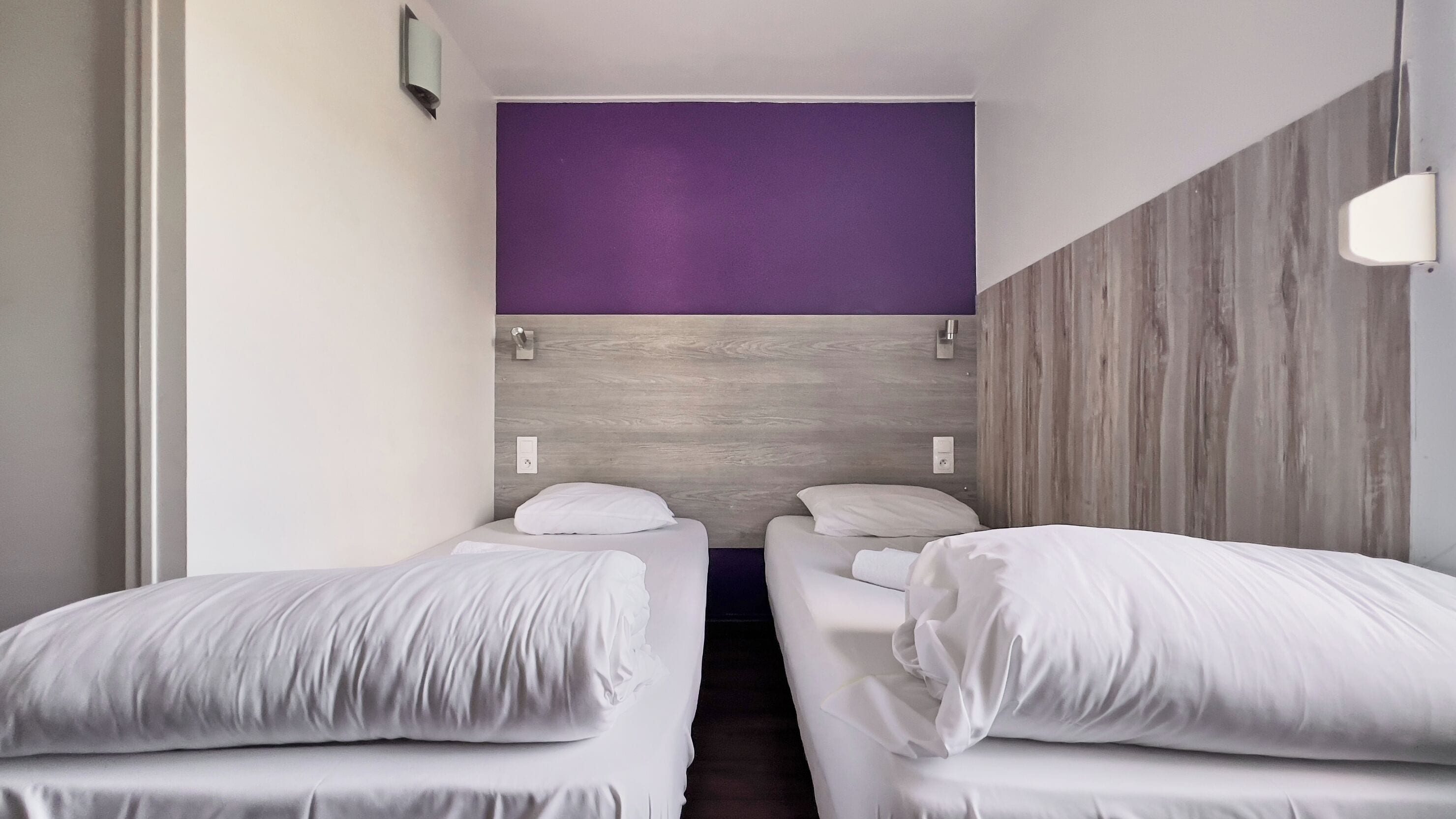 Quarto de hotel com duas camas de solteiro, uma parede de destaque roxa e uma cabeceira de madeira.
